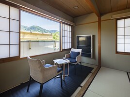 Quarto com piso de tatame, uma mesa baixa, duas cadeiras e uma porta deslizante que dá para uma varanda com vista para uma montanha e uma cerca de madeira.
