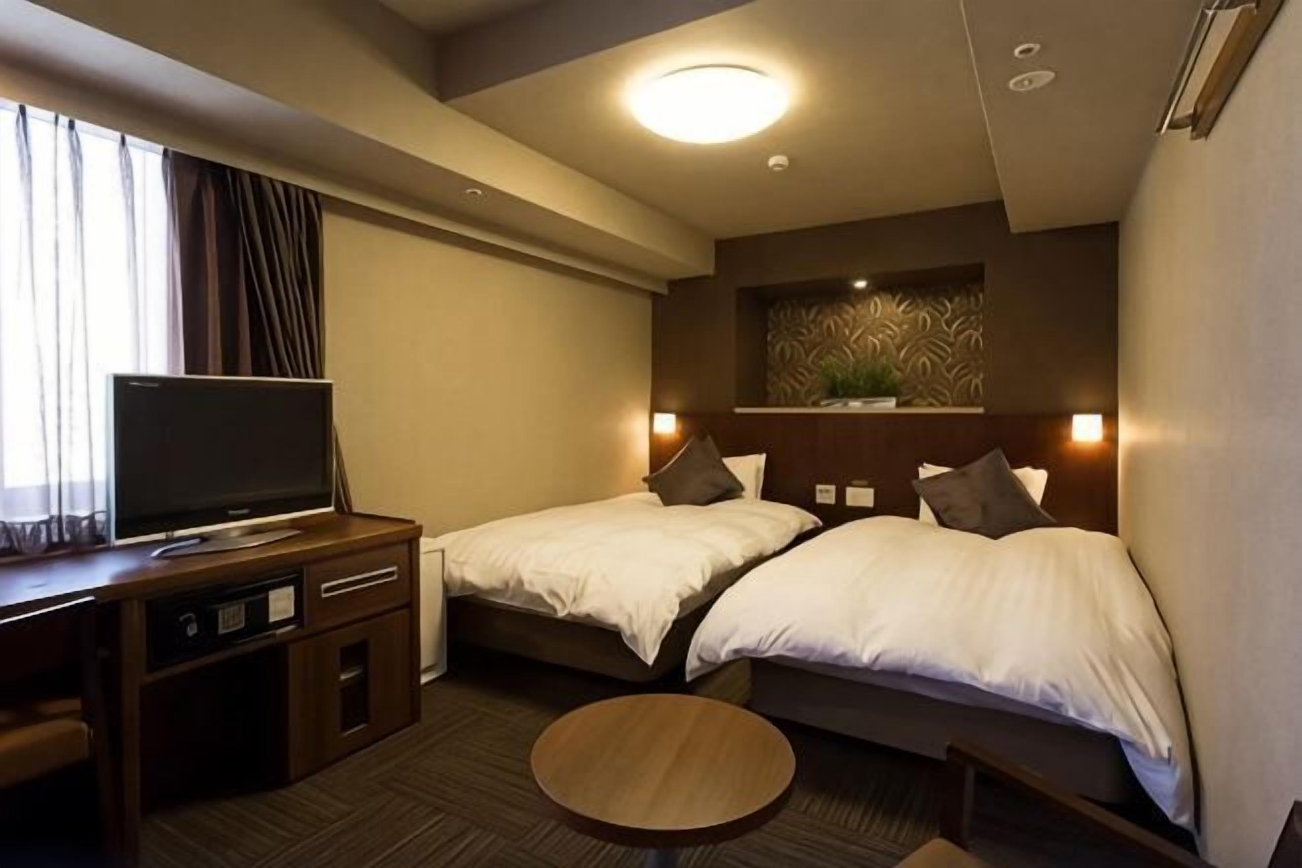 Quarto de hotel com duas camas, uma escrivaninha com televisão e uma mesinha redonda.