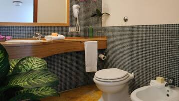 Casa de banho com sanita, bidé e lavatório. Tem um porta-toalhas com uma toalha, um secador de cabelo e um espelho sobre o lavatório.