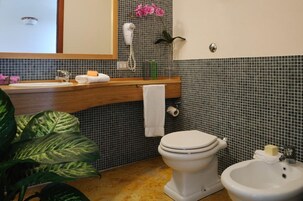 Casa de banho com sanita, bidé e lavatório. Tem um porta-toalhas com uma toalha, um secador de cabelo e um espelho sobre o lavatório.