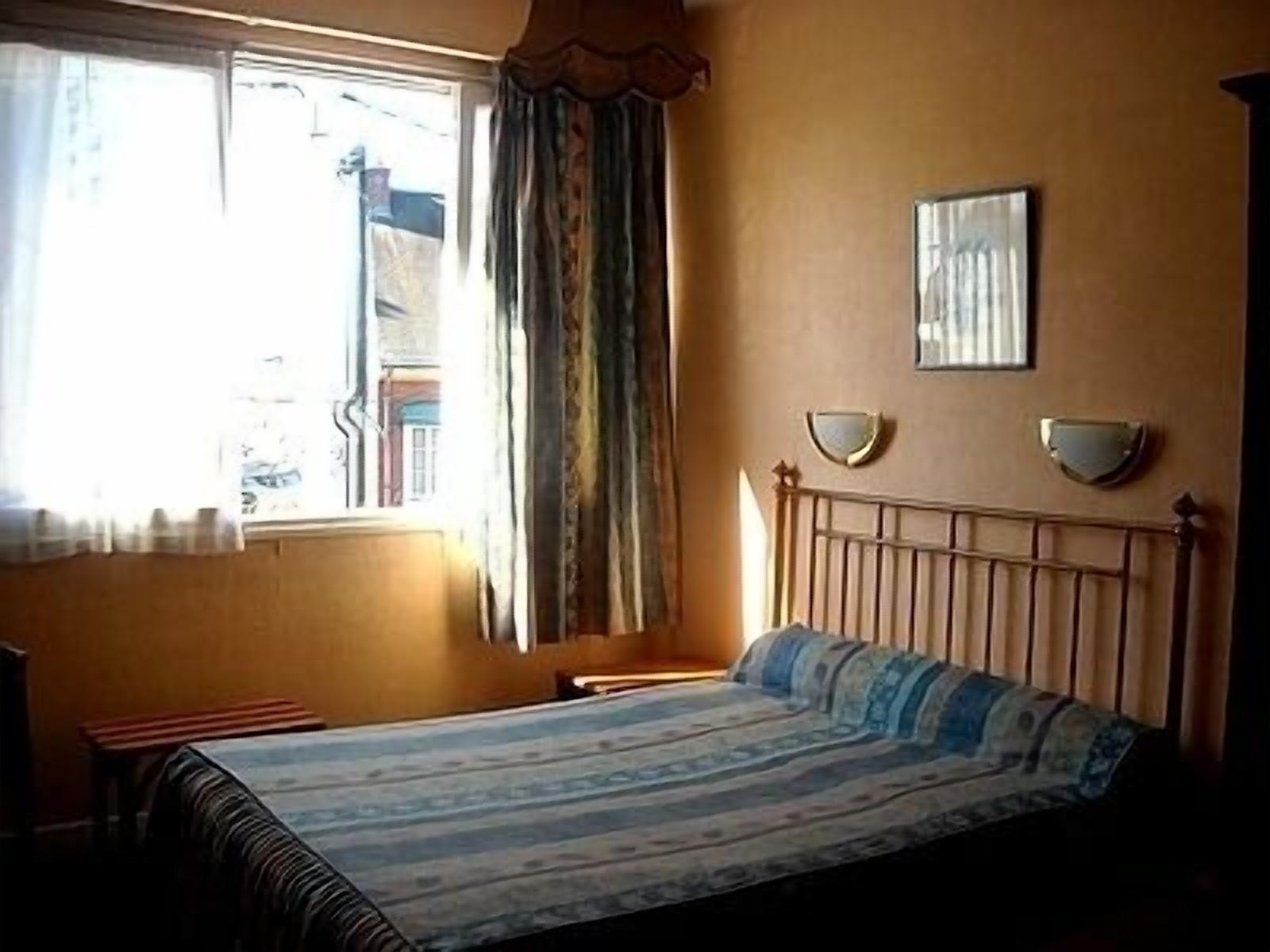 Quarto com cama, janela com cortinas e luminária fixada na parede.
