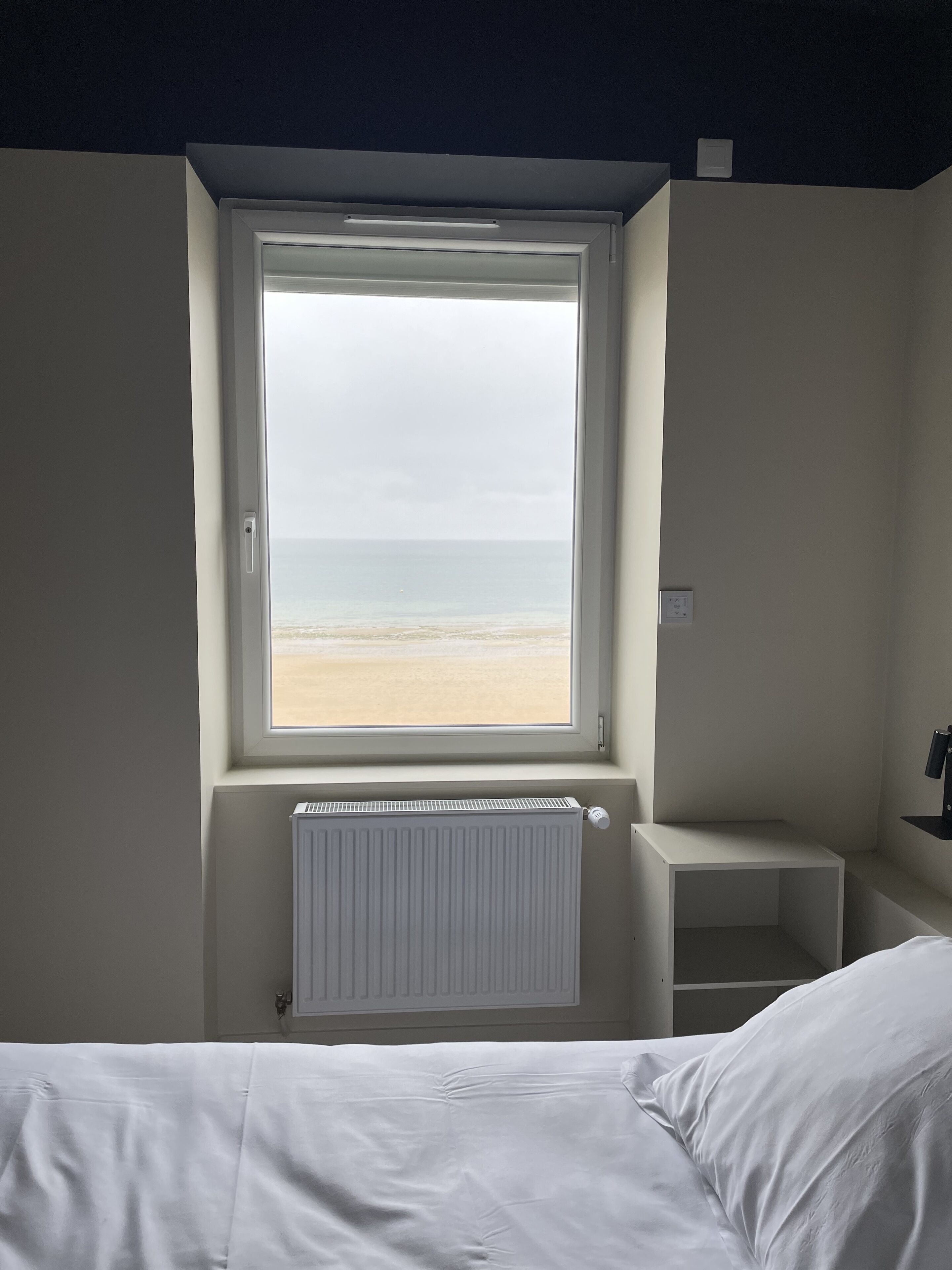 Quarto com uma grande janela com vista para uma praia, um radiador e uma mesinha de cabeceira.