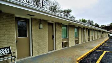 Um motel com uma fileira de quartos térreos, um banco na parte externa e estacionamento com vagas demarcadas.