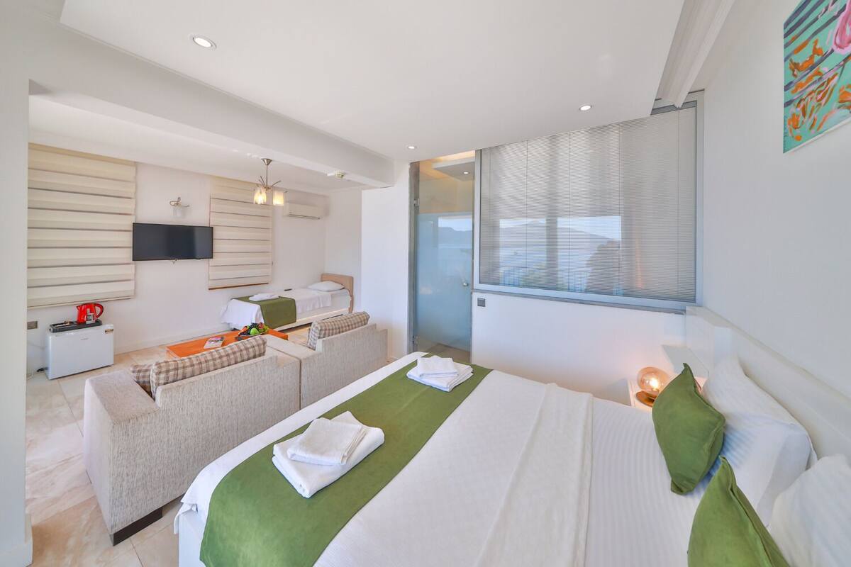 Quarto de hotel moderno com cama grande, sofá, televisão e janela com persianas.