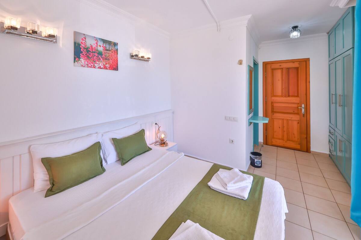 Quarto com cama, almofadas, mesa de cabeceira com abajur, pintura na parede e porta de madeira.