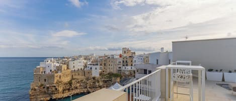 Uma vila costeira com edifícios ao longo do penhasco, uma varanda com mesa e cadeiras, e vista para o mar.