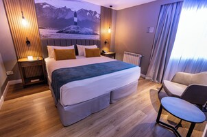 Quarto de hotel com uma cama grande, duas mesas de cabeceira, uma cadeira, televisão fixada na parede e uma janela com cortinas.