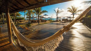 Uma rede sob um deck de madeira, com vista para uma praia e palmeiras.