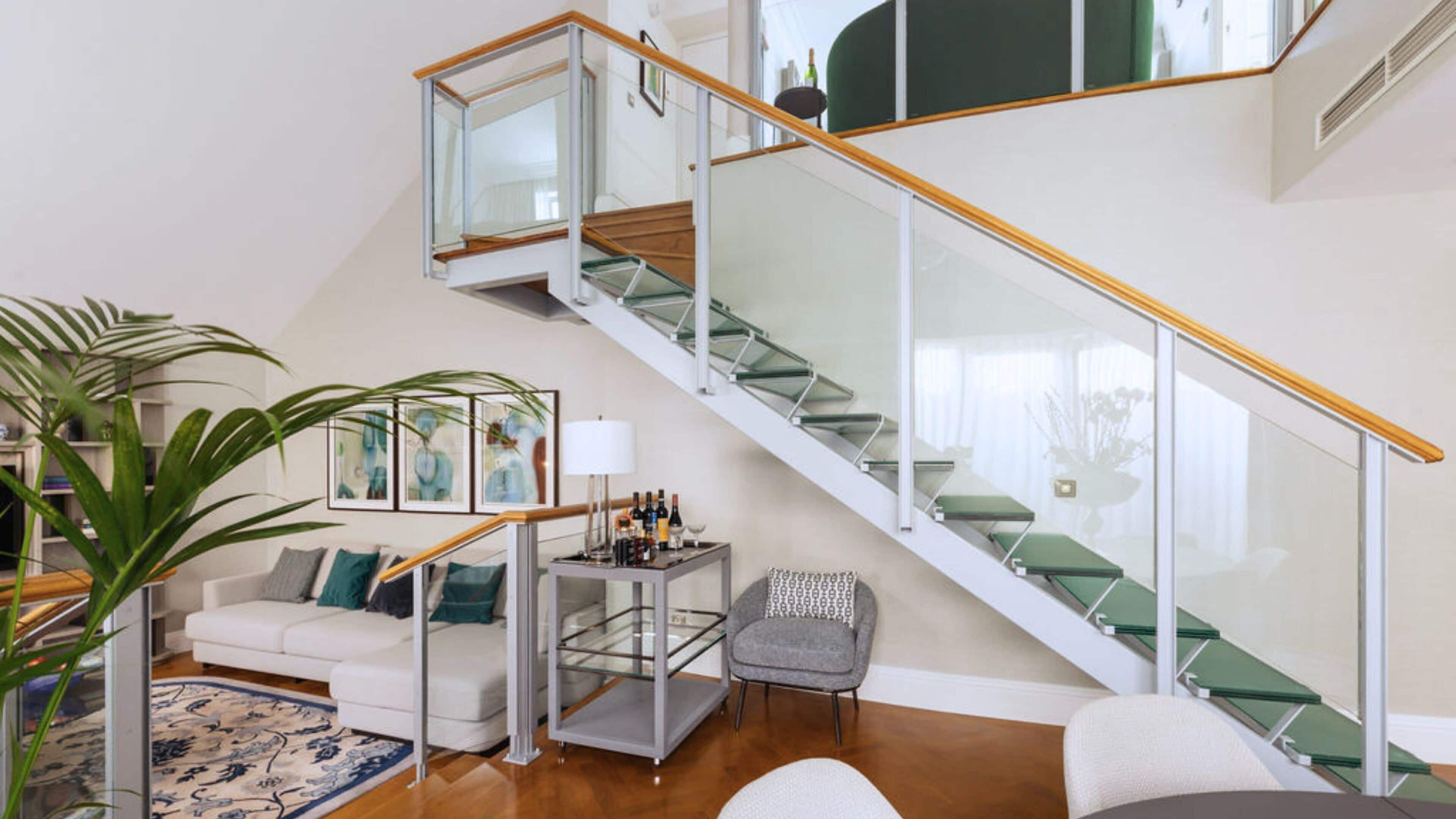 Uma sala de estar moderna com escada de vidro, sofá branco, mesinha com bebidas e poltrona verde.