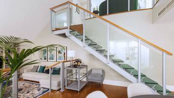 Uma sala de estar moderna com escada de vidro, sofá branco, mesinha com bebidas e poltrona verde.