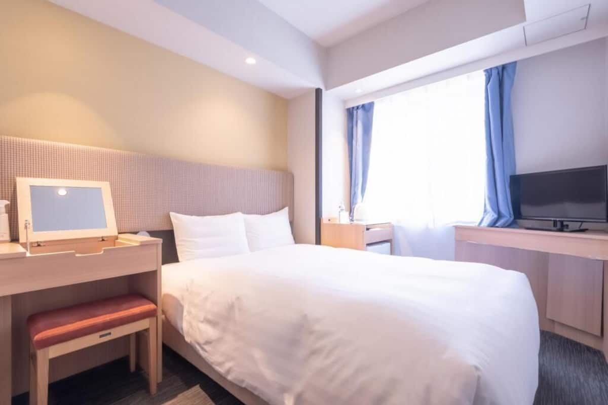 Quarto de hotel com cama, escrivaninha, televisão e janela com cortinas.