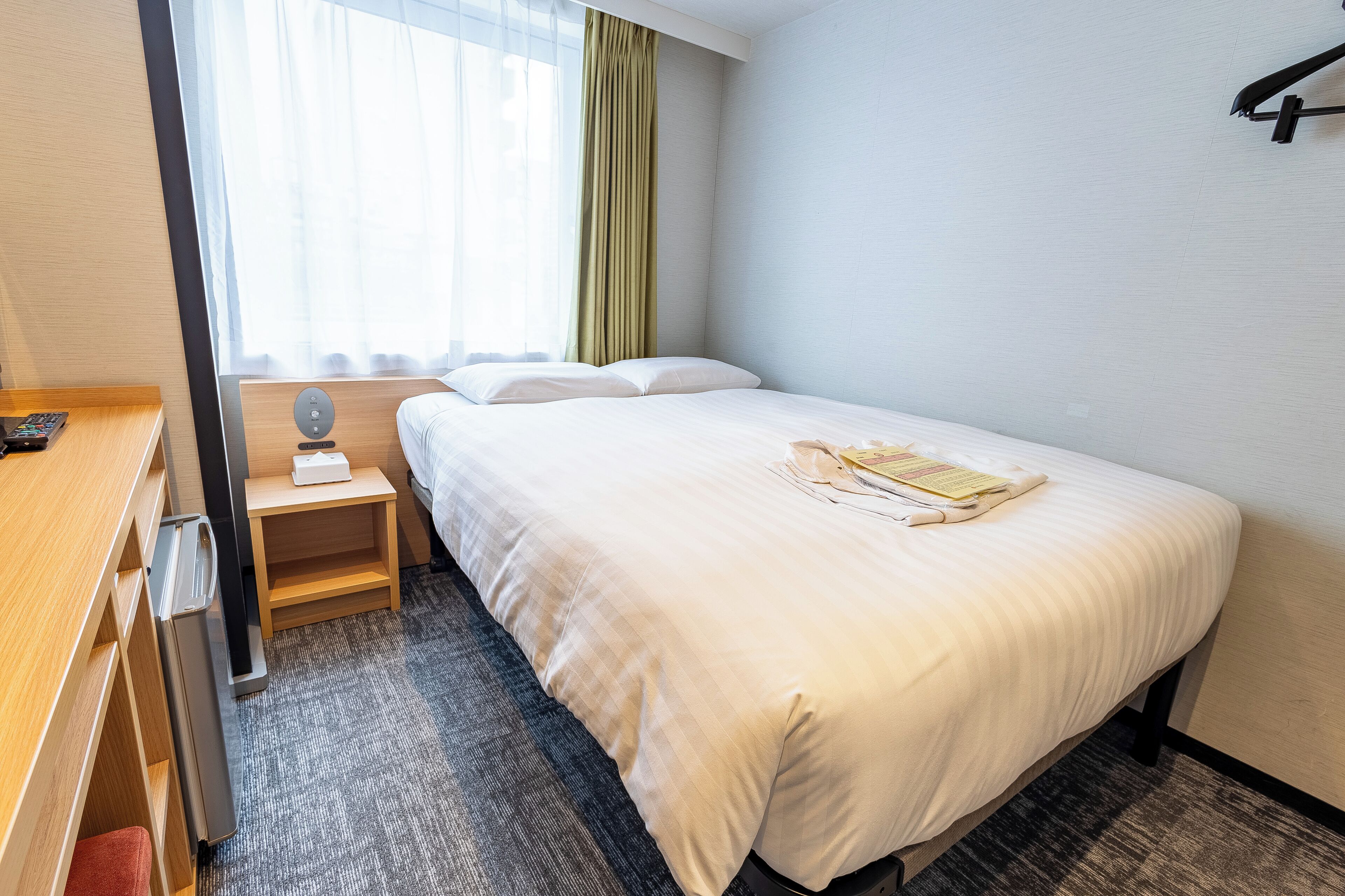 Quarto de hotel com cama, escrivaninha, mesa de cabeceira, abajur e janela com cortinas.