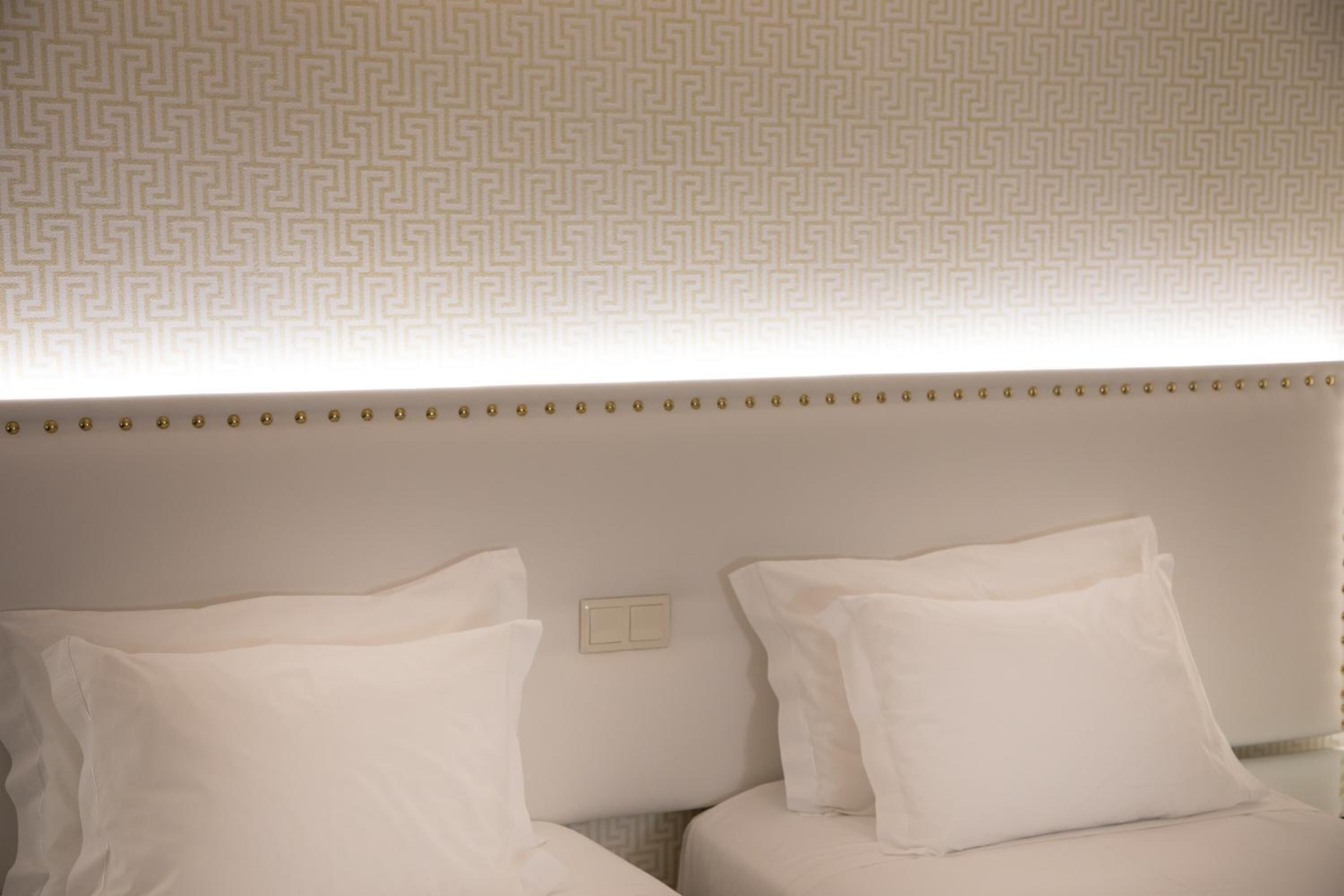 Quarto de hotel com papel de parede estampado, cabeceira branca com detalhes dourados e duas almofadas brancas.