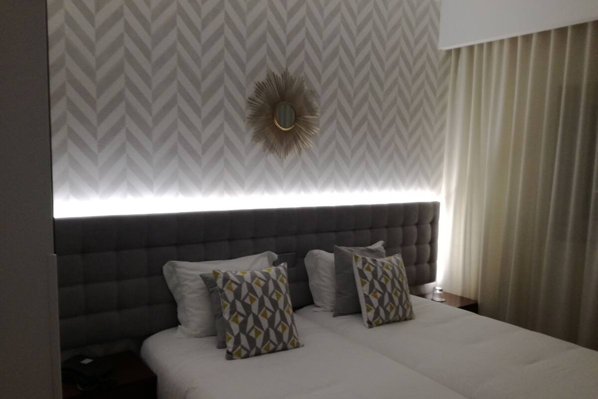 Quarto de hotel com uma cama grande, cabeceira estofada e cortinas estampadas.