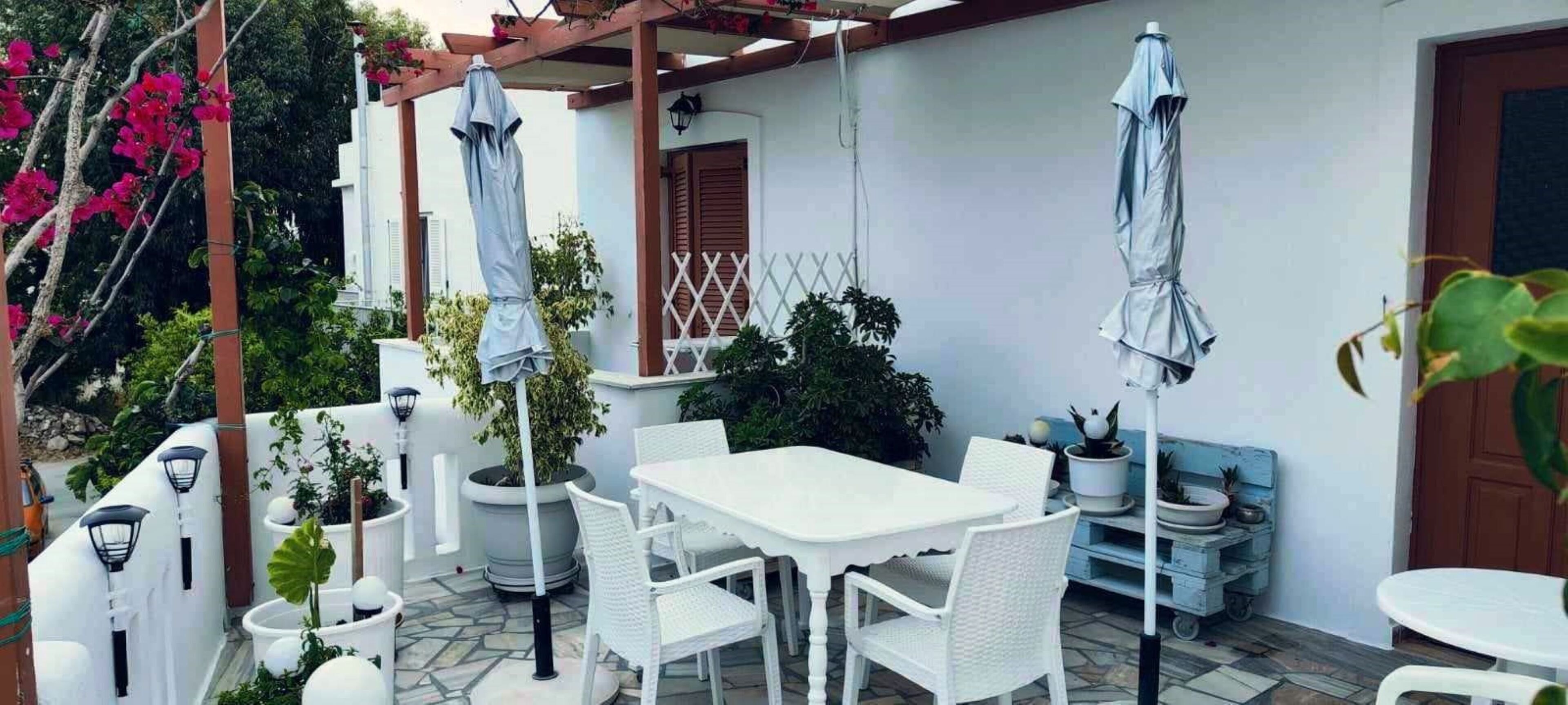 Um terraço ao ar livre com mesas e cadeiras brancas, guarda-sóis, plantas em vasos e uma pérgula.