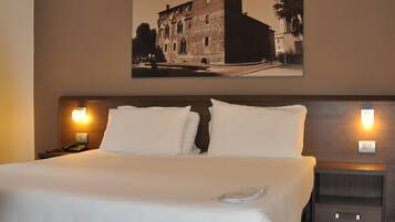 Quarto de hotel com uma cama grande, uma mesinha de cabeceira, uma luminária na parede e um quadro com a imagem de um edifício.