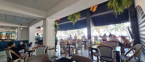 Uma área de estar interna com cadeiras e mesas de vime, um bar ao fundo e vista para o exterior.