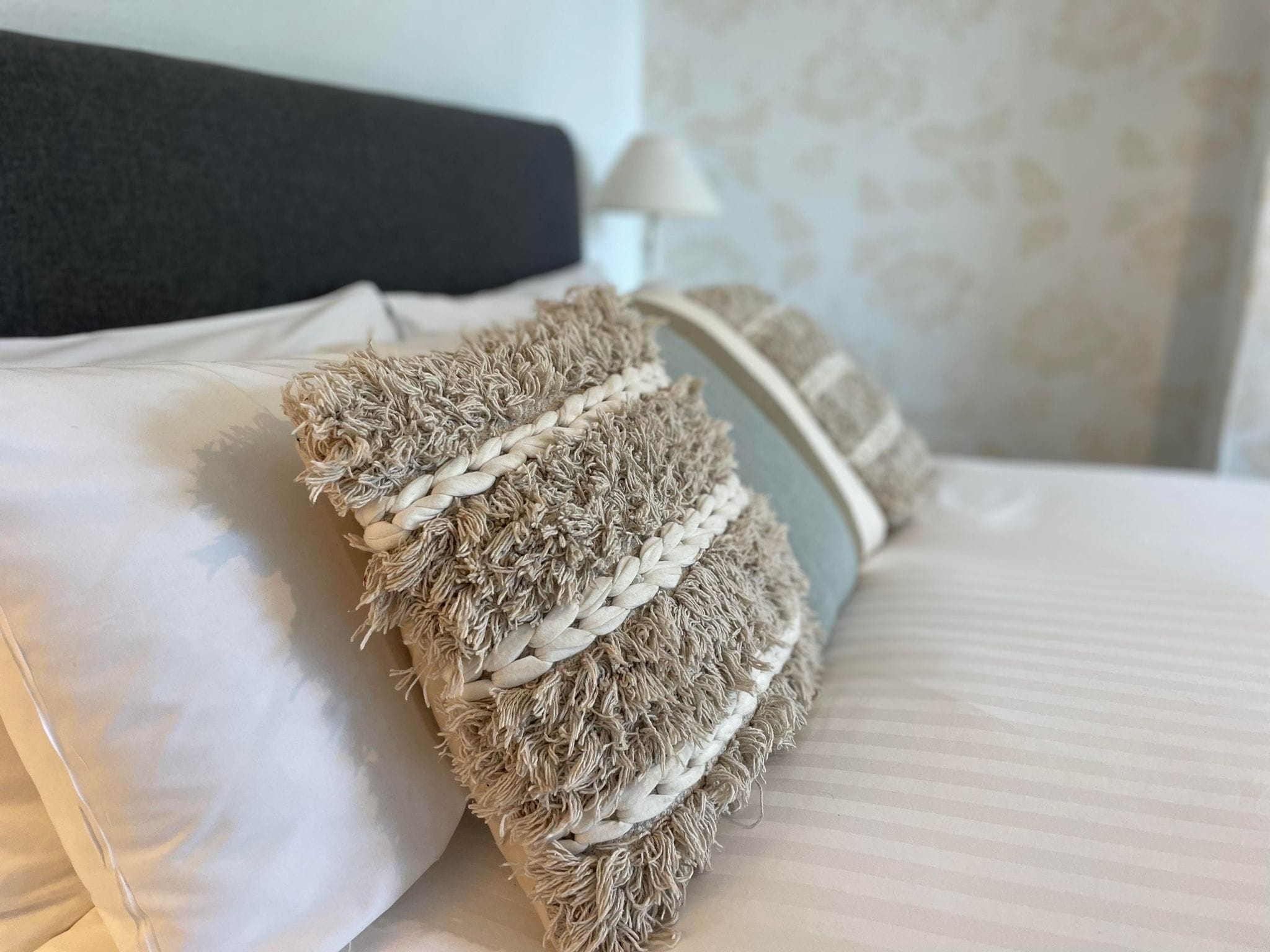 Uma cama com roupa de cama branca e um travesseiro bege texturizado.