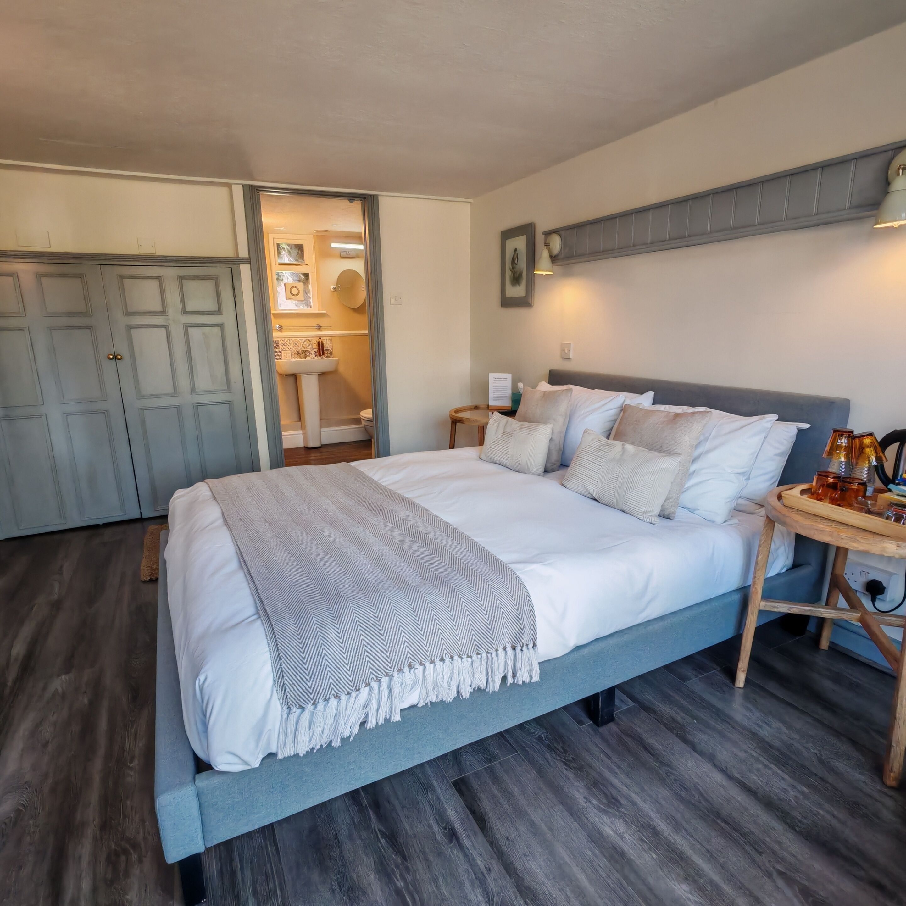 Quarto com cama grande, mesa de cabeceira, armário e banheiro visível por uma porta aberta, além de piso de madeira.