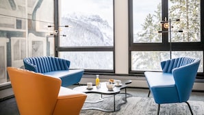 Quarto moderno com duas poltronas azuis, uma mesa de centro pequena e vista para montanhas cobertas de neve através de grandes janelas.