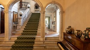 Uma escadaria imponente com carpete verde, ladeada por colunas e arcos ornamentados, conduzindo a um piano e a um cômodo com um espelho grande e arranjo floral.