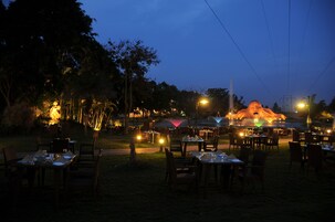 Uma área de refeições ao ar livre com mesas e cadeiras, cercada por árvores e iluminada por iluminação ambiente.