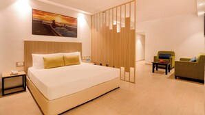 Quarto de hotel com uma cama grande, um separador de madeira, uma poltrona verde e uma pequena mesa.