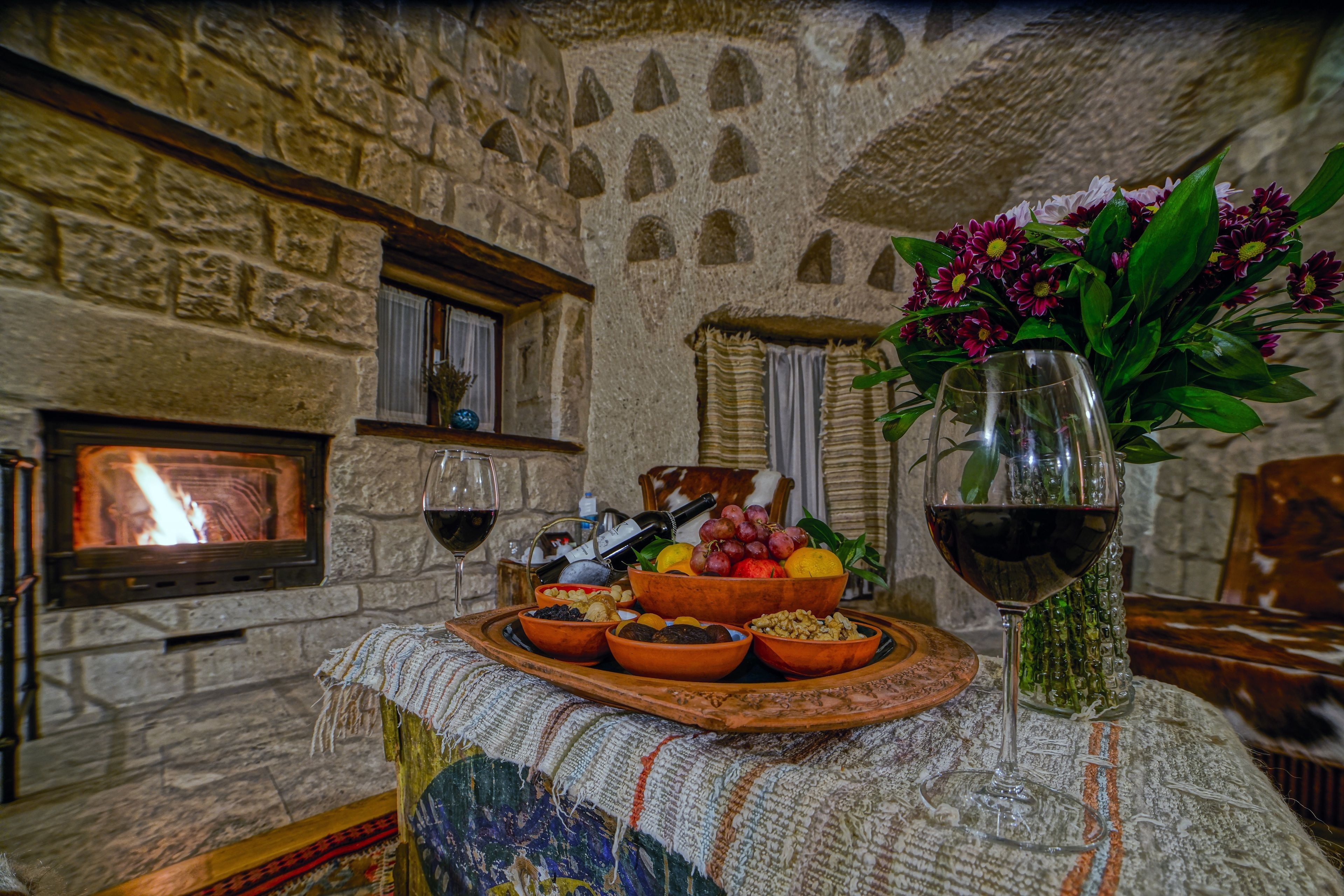 Quarto com paredes de pedra, lareira, uma mesa com uma fruteira, uma taça de vinho e um vaso com flores.