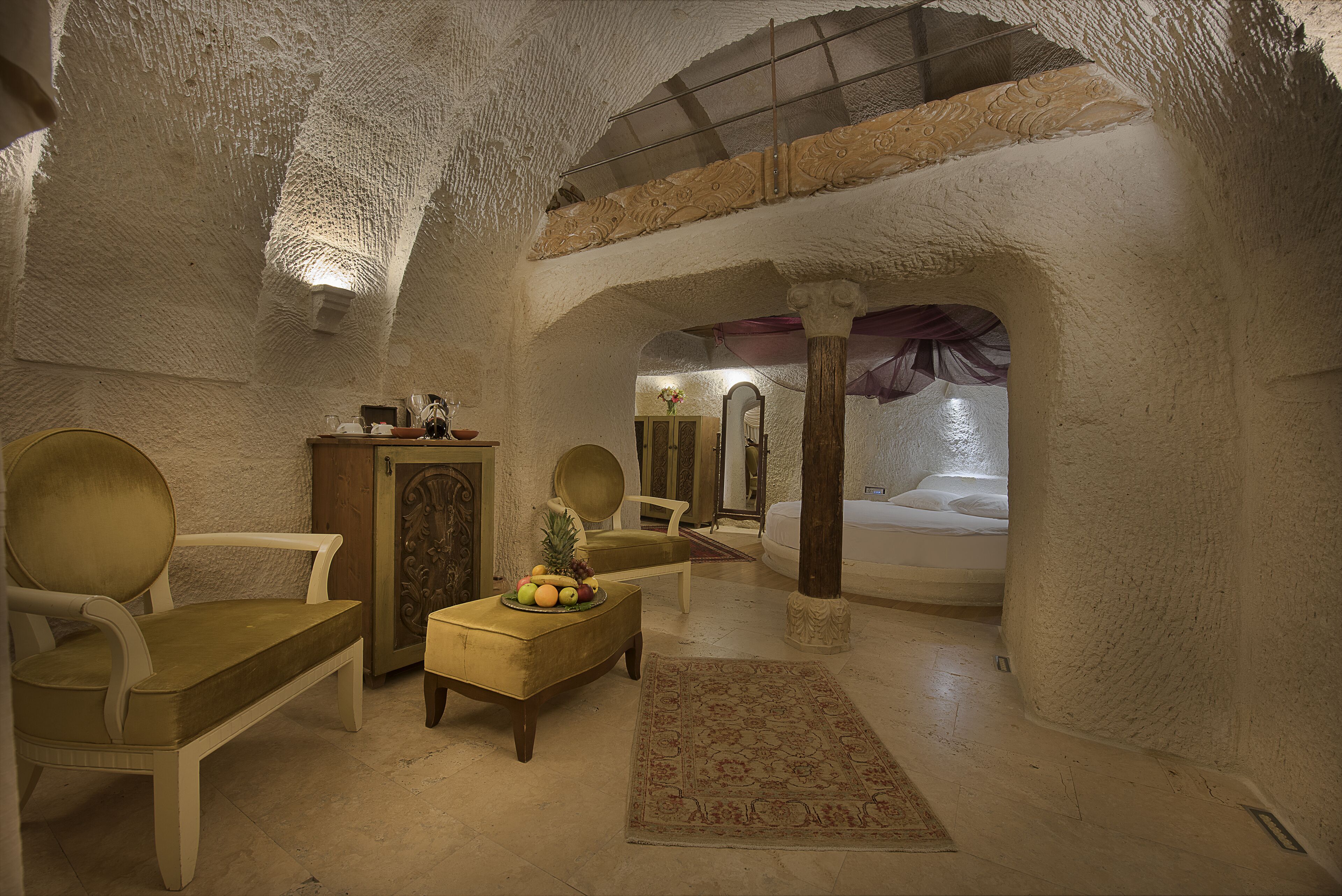 Um quarto com interior singular em estilo de caverna, com uma cama, duas cadeiras, uma mesa pequena com frutas e um tapete decorativo.