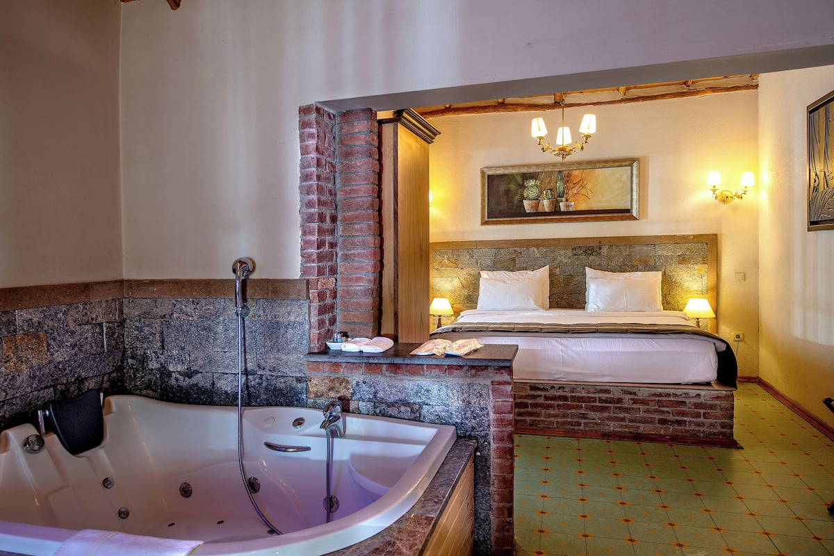 Quarto com uma banheira grande, cama com duas almofadas, mesa pequena com abajur e um quadro na parede.