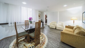 Uma sala de estar moderna com uma mesa de jantar de vidro, duas cadeiras de madeira, um sofá bege, um tapete estampado e um abajur sobre uma mesa lateral.