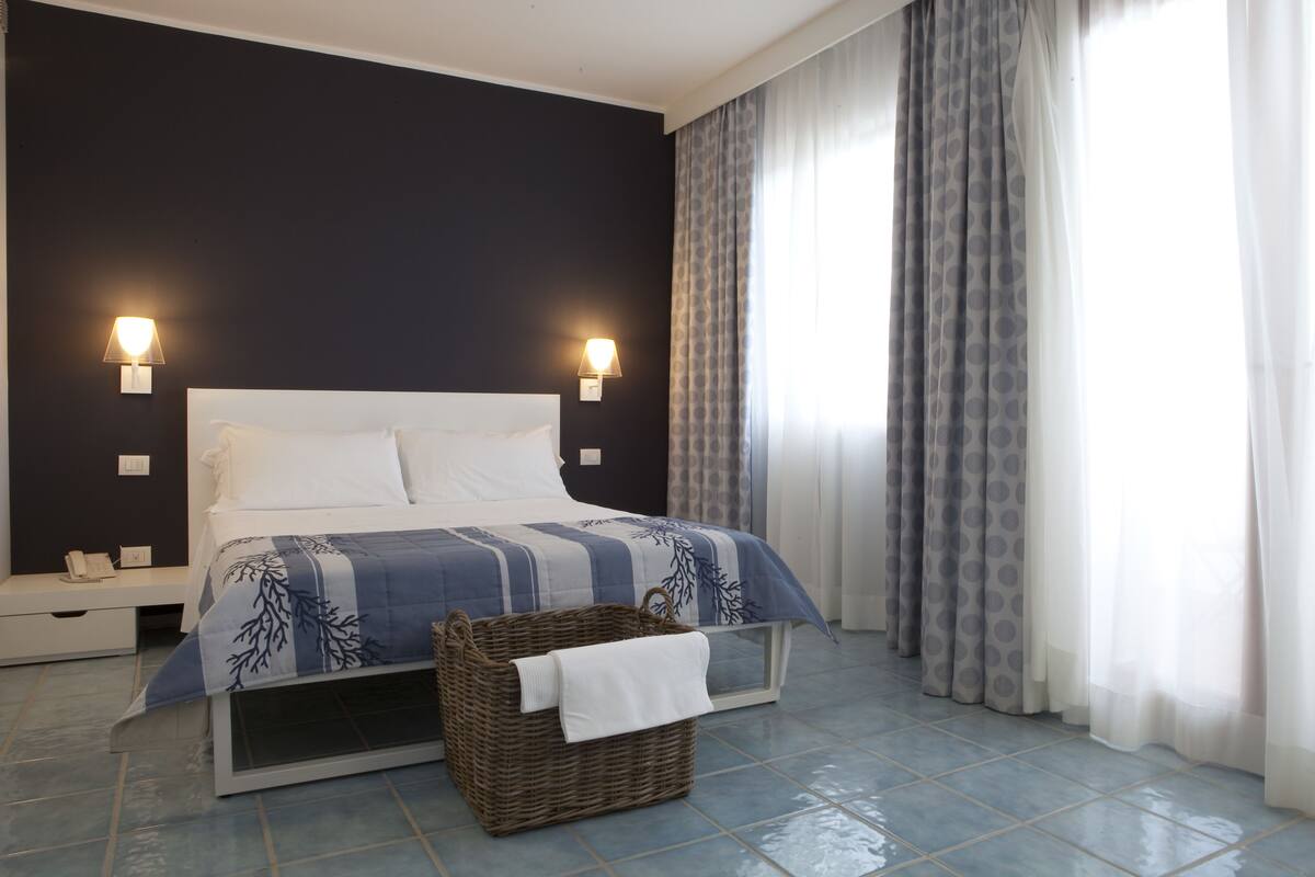 Quarto com cama, duas luminárias de cabeceira, mesa de cabeceira, cesto de vime e janela com cortinas.
