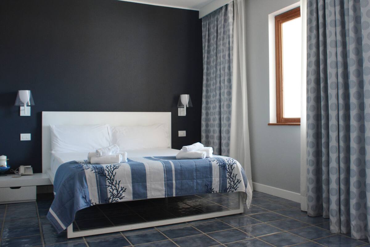Quarto com cama, duas luminárias de cabeceira, janela com cortinas e mesa de cabeceira com abajur.