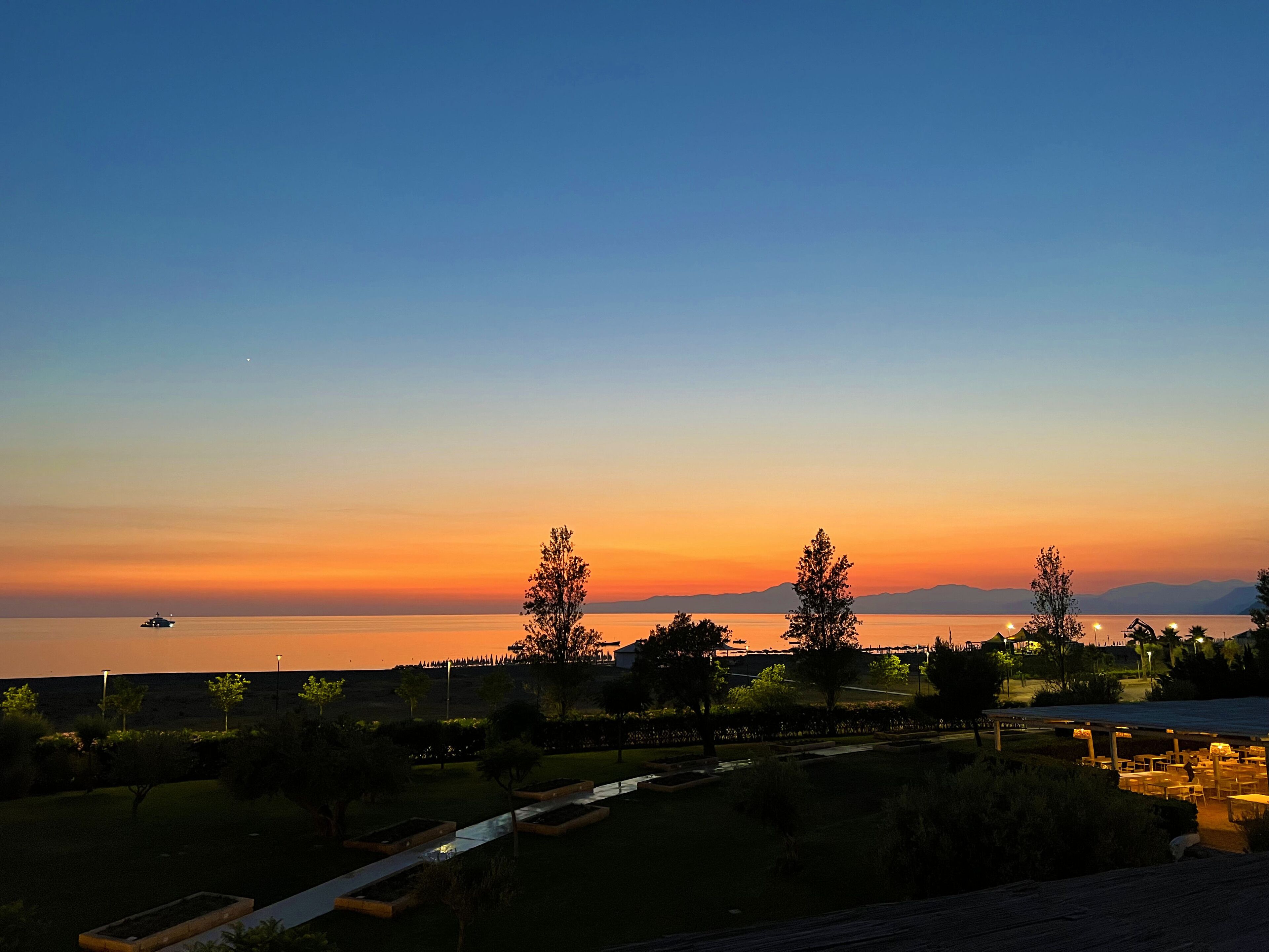 Vista do pôr do sol sobre um lago calmo, com um barco, árvores e uma área de estar ao ar livre bem iluminada.