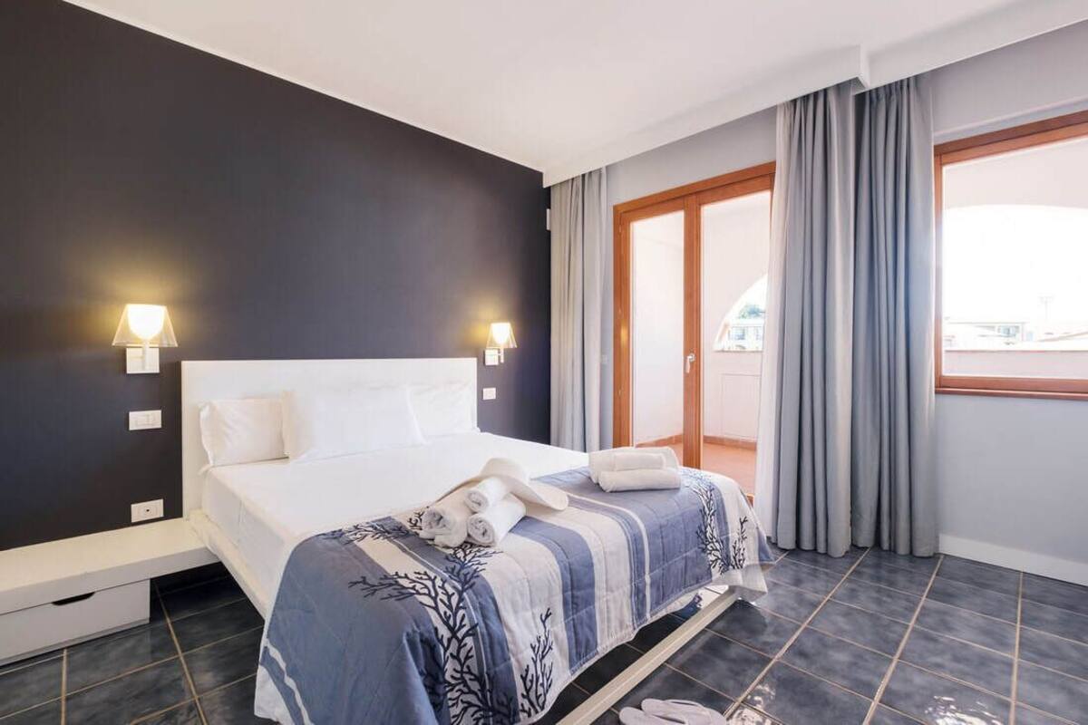 Quarto de hotel com cama, duas luminárias fixadas na parede, uma janela grande com cortinas e vista para o exterior.