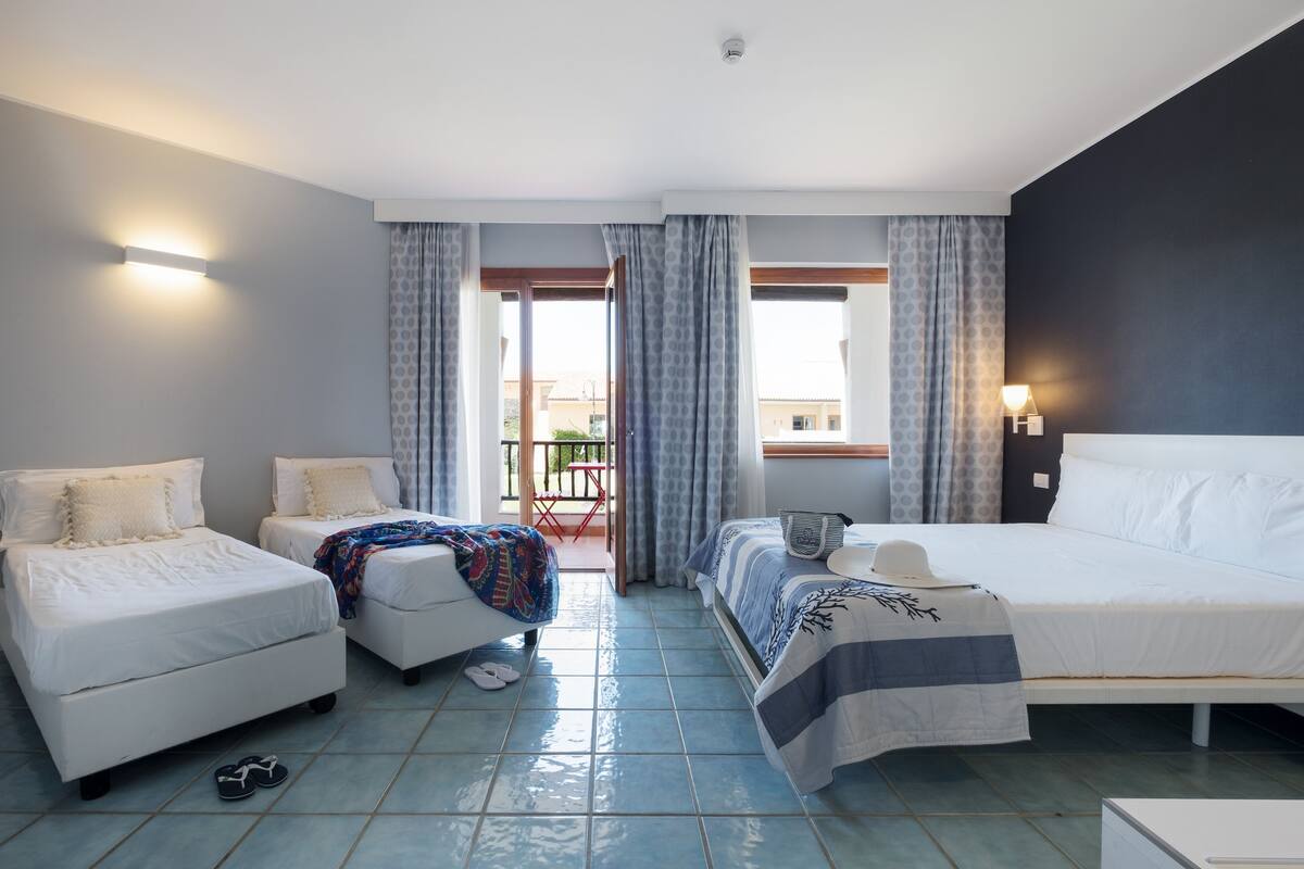 Quarto de hotel com duas camas de solteiro, um pequeno sofá, piso ladrilhado e varanda com cortinas.