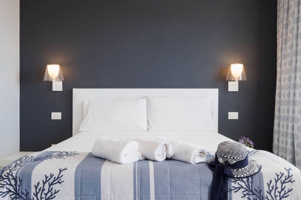 Uma cama bem arrumada com lençóis brancos e manta listrada, duas luminárias fixadas na parede e um chapéu estampado sobre a cama.