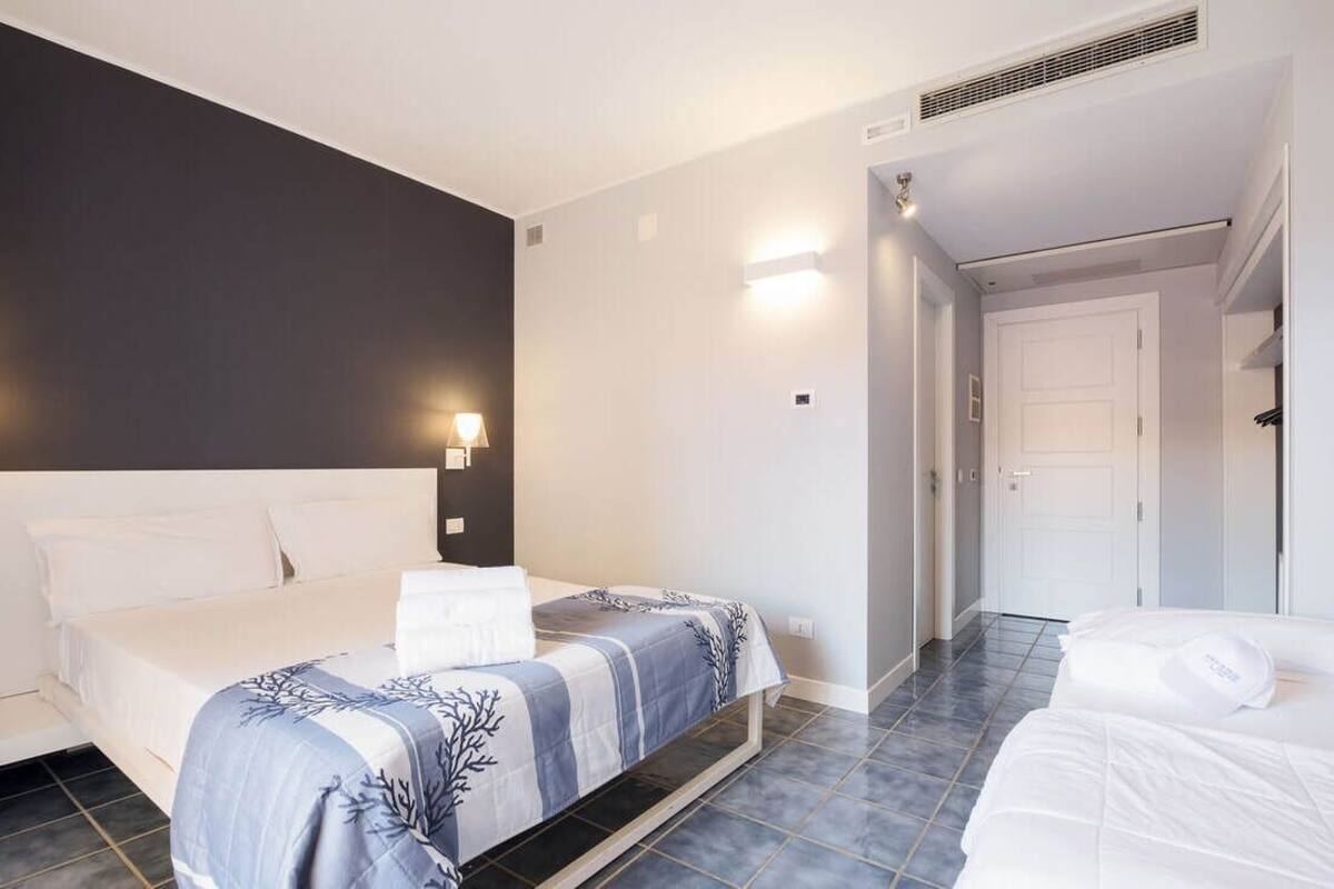 Quarto de hotel com cama, mesa de cabeceira, luminária na parede e porta para outro cômodo.