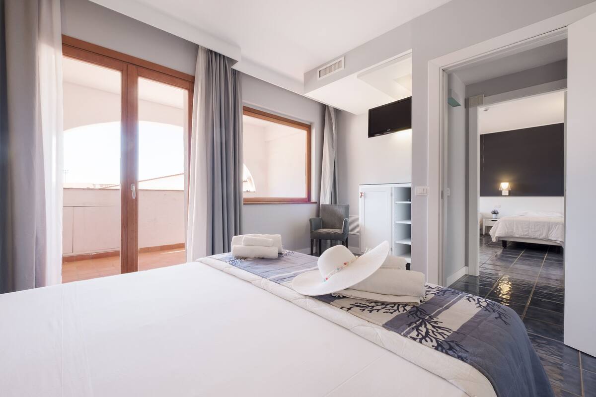 Quarto de hotel moderno com uma cama grande, área de estar com uma poltrona, escrivaninha e vista para a varanda.
