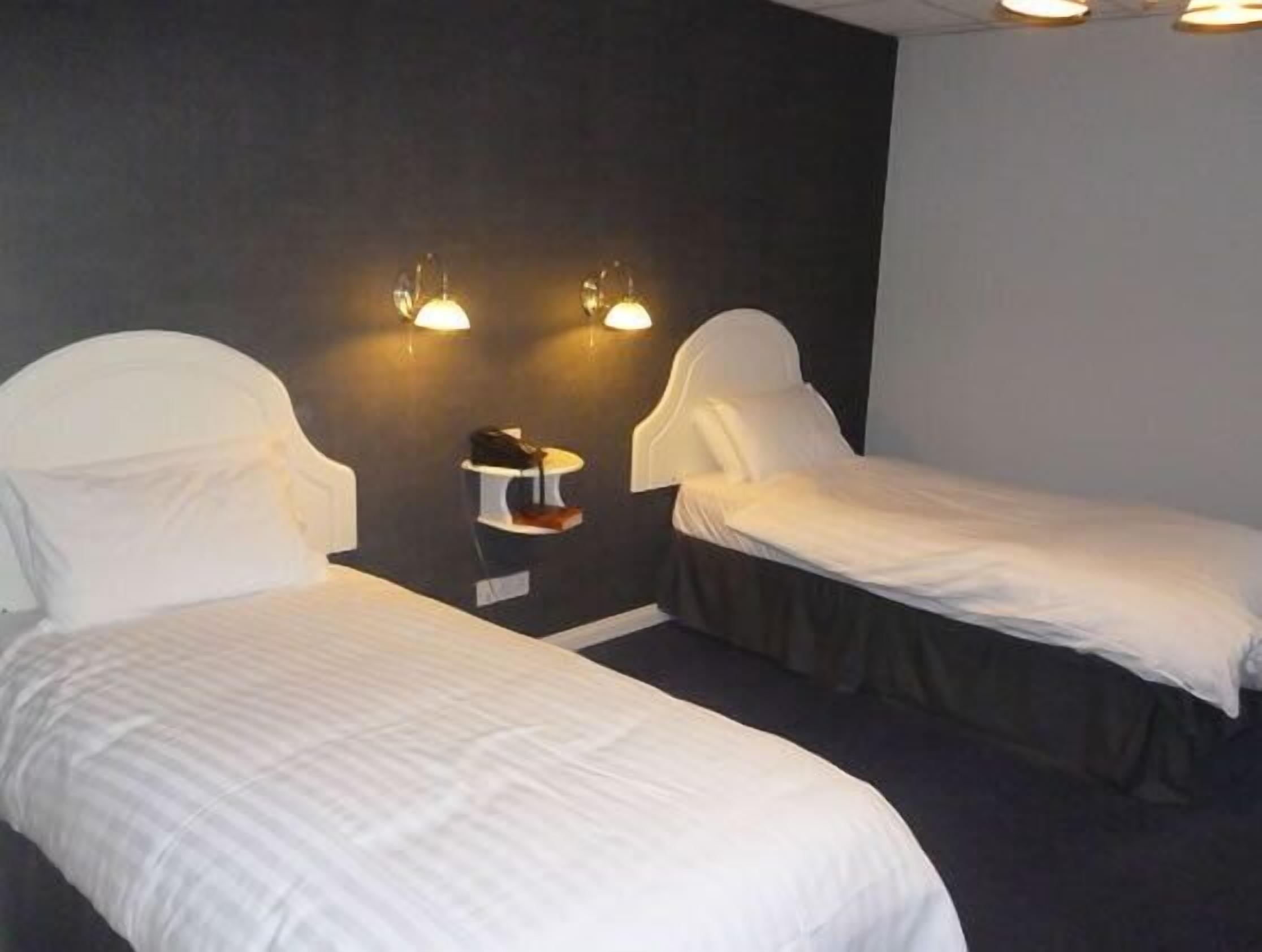 Quarto de hotel com duas camas de solteiro, uma pequena mesa de cabeceira com abajur e luminária na parede.