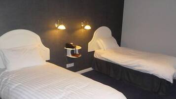 Quarto de hotel com duas camas de solteiro, uma pequena mesa de cabeceira com abajur e luminária na parede.