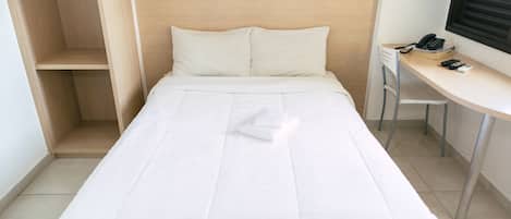 Uma cama bem arrumada com roupa de cama branca, cabeceira de madeira, uma escrivaninha com telefone e uma cadeira.