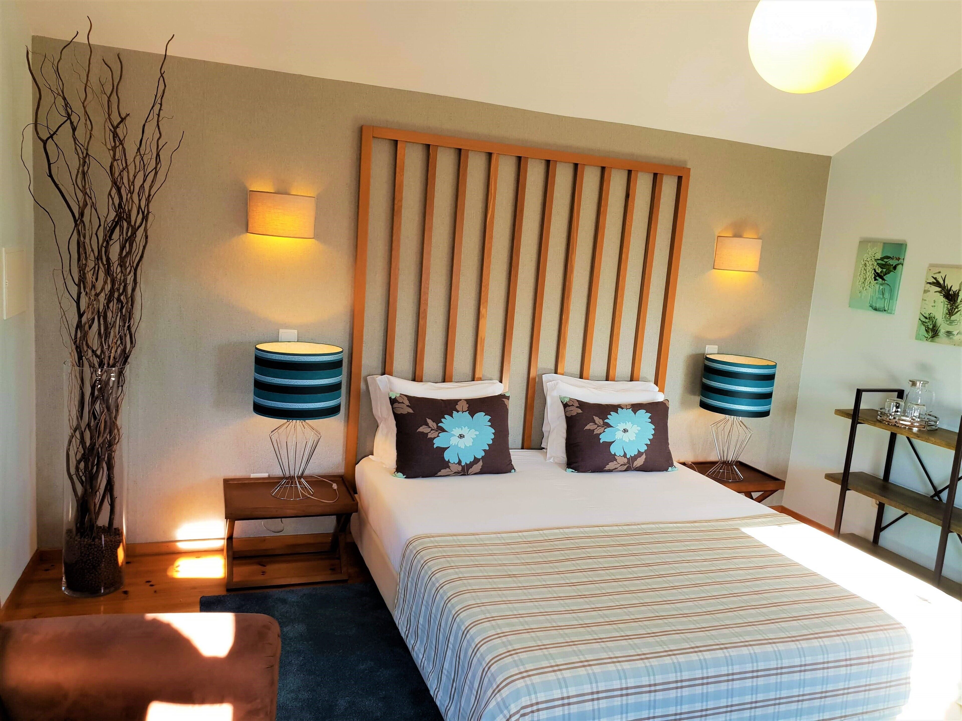 Quarto com cama, duas luminárias de cabeceira, mesa de apoio, um vaso com galhos e uma pequena mesa com objetos decorativos.