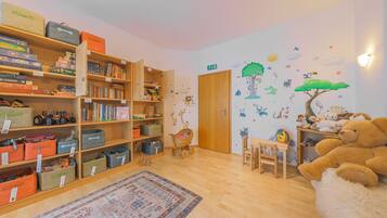 Quarto infantil com prateleiras de madeira repletas de livros e brinquedos, berço de madeira, um urso de pelúcia e uma parede decorada com adesivos de animais.