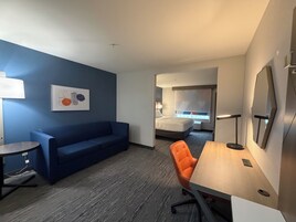 Quarto de hotel com sofá azul, cadeira laranja, mesa de madeira, cama com cabeceira branca e um espelho grande.