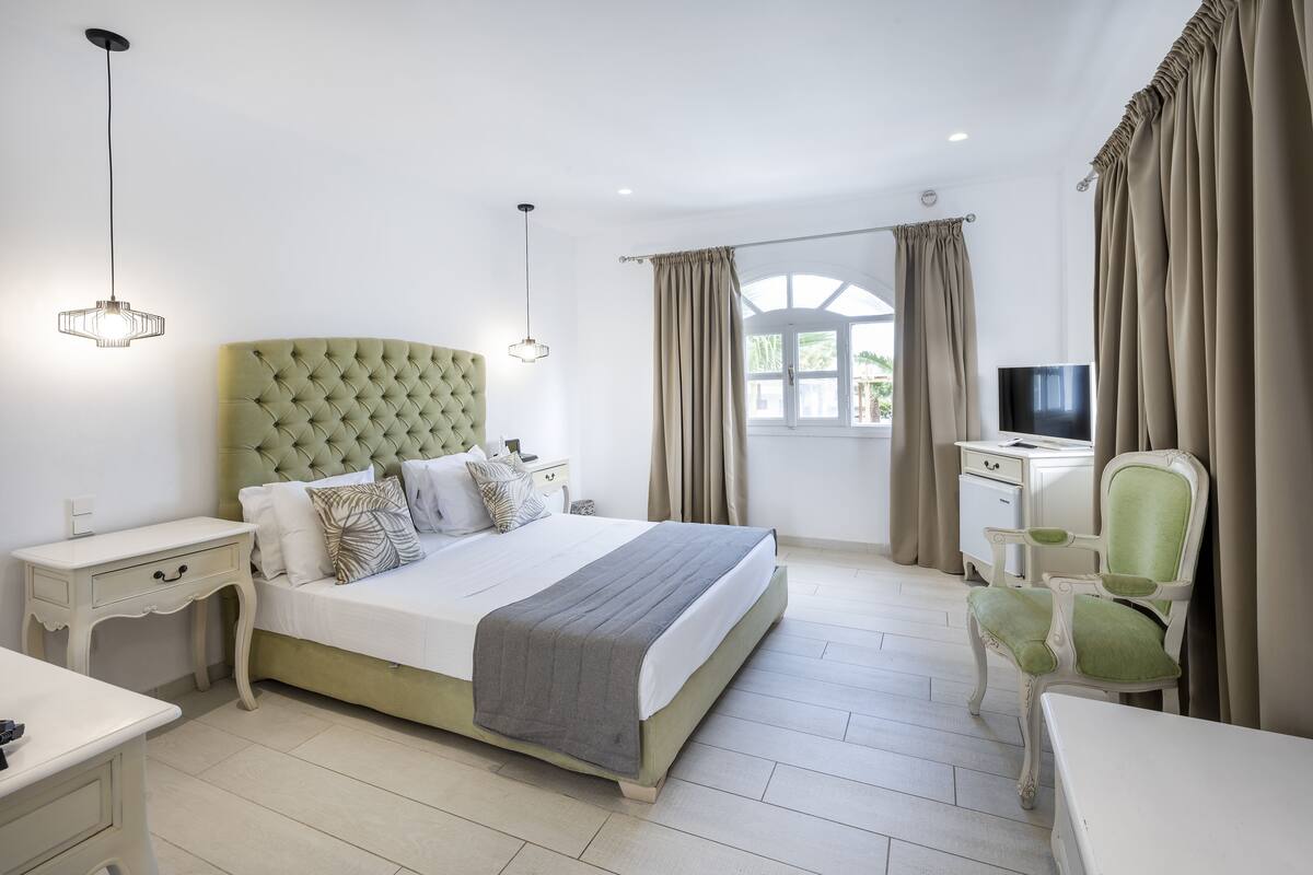 Quarto com cabeceira estofada, cama com cobertor cinza, mesa de cabeceira, escrivaninha com cadeira e televisão.