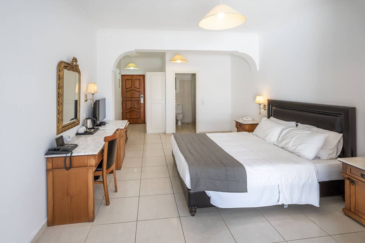 Quarto de hotel com cama, escrivaninha, cadeira e espelho.