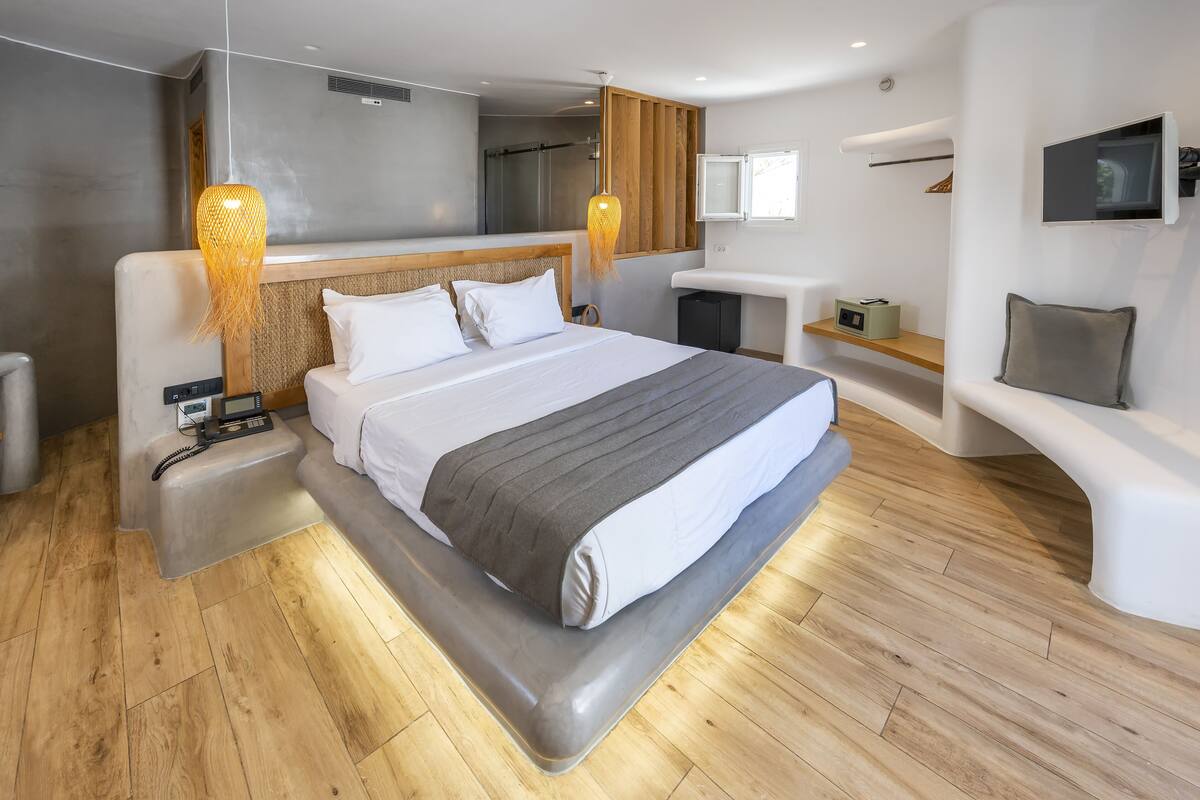 Quarto de hotel moderno com uma cama grande, mesas de cabeceira, televisão e área de chuveiro.