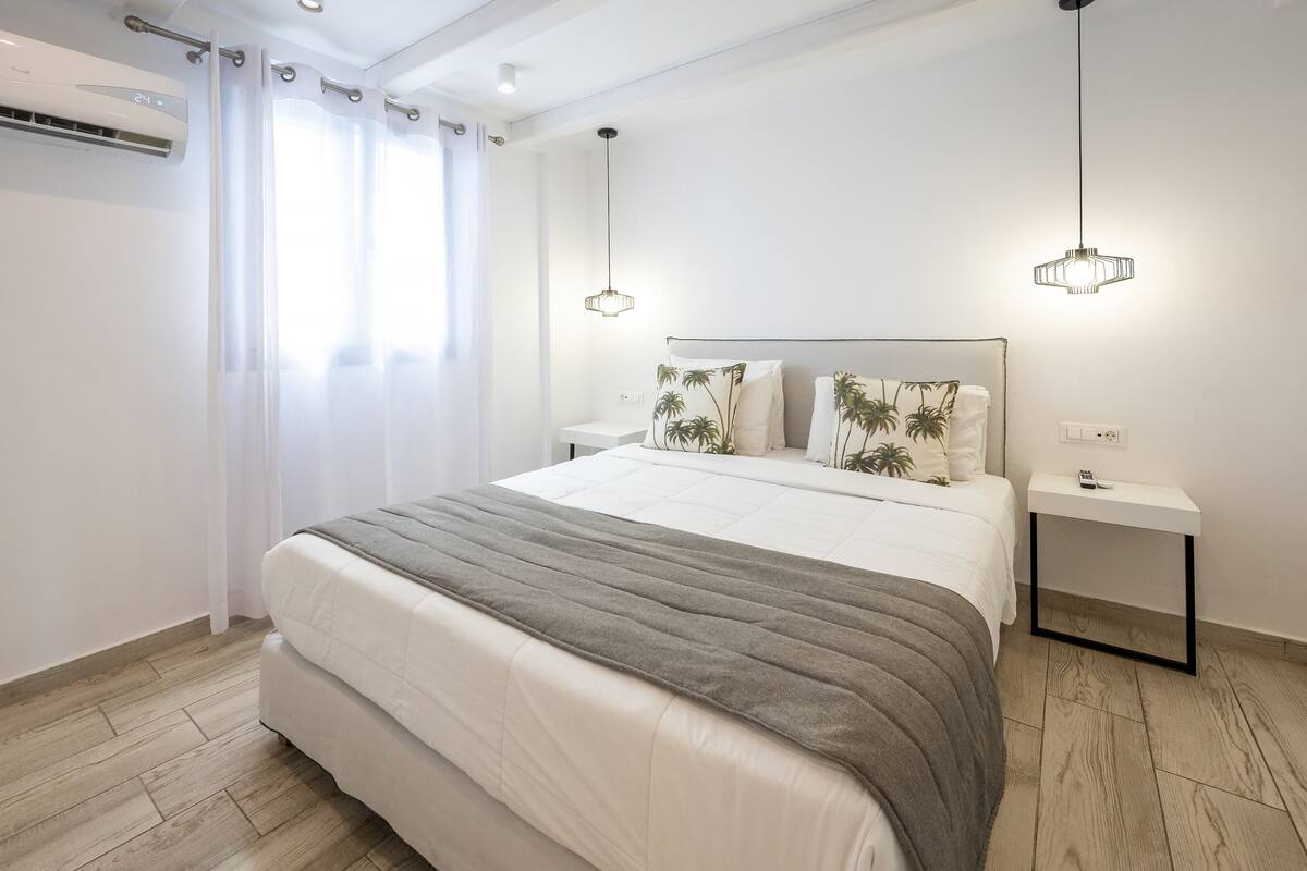 Um quarto moderno com uma cama grande, duas mesas de cabeceira, um criado-mudo, um lustre e uma janela com cortinas.