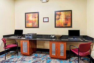 Quarto de hotel com escrivaninha, duas cadeiras, dois laptops, telefone e duas pinturas abstratas emolduradas na parede.